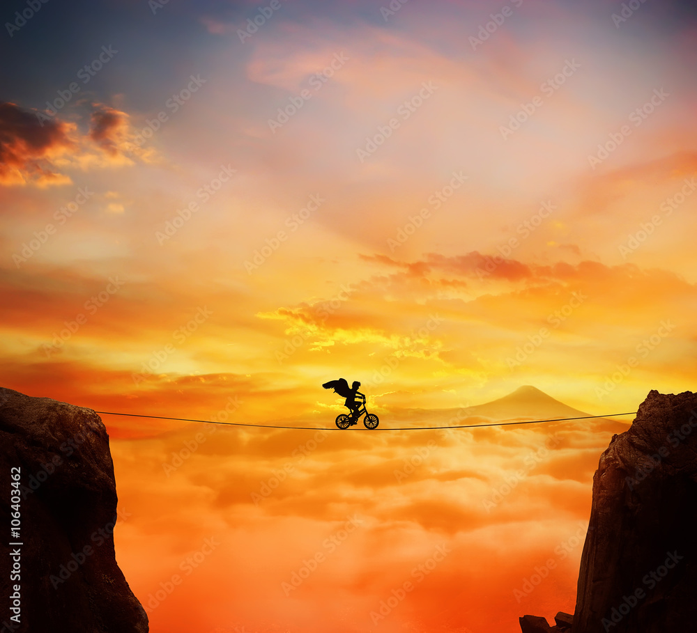 Boy with angel wings balance on a rope over a chasm riding a bicycle ...