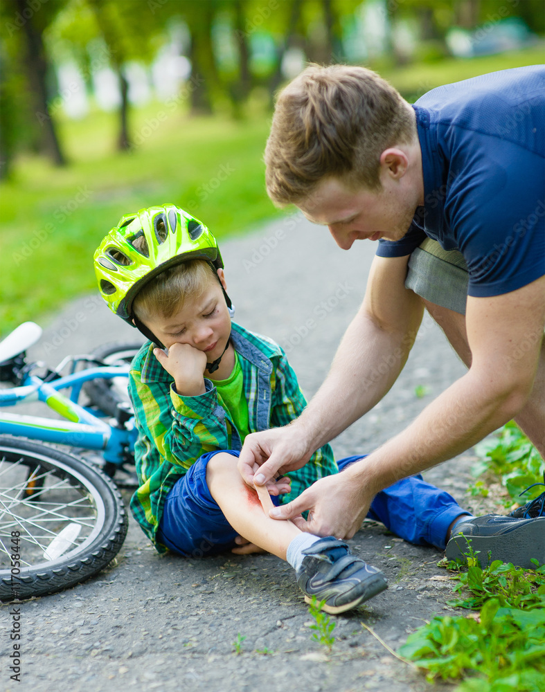 father putting band-aid on young boy's injury who fell off his bike ...
