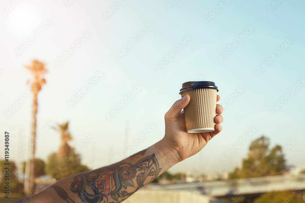 Arm and hand of a tattooed man with a beige plain coffee cup against ...