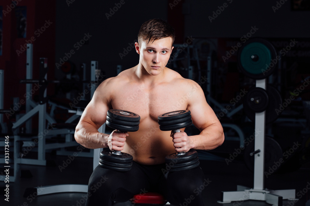professional bodybuilder sitting on the bench, resting between exercises with dumbbells at gym ...