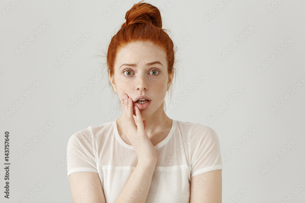 Frightened startled young redhead freckled female raising eyebrows, keeping hand on her cheek ...