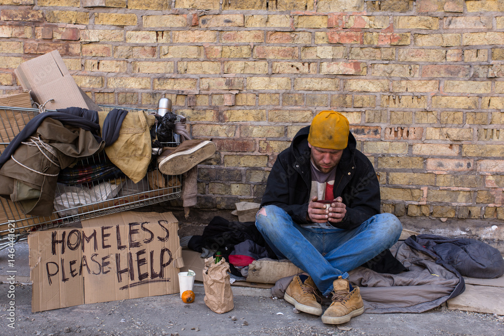 Homeless man sitting on the street, near the wall and cart with his ...