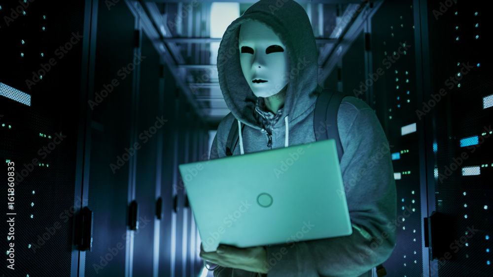 Close-up Shot of a Masked Hacker in a Hoodie Standing in the Middle of Data Center full of Rack ...