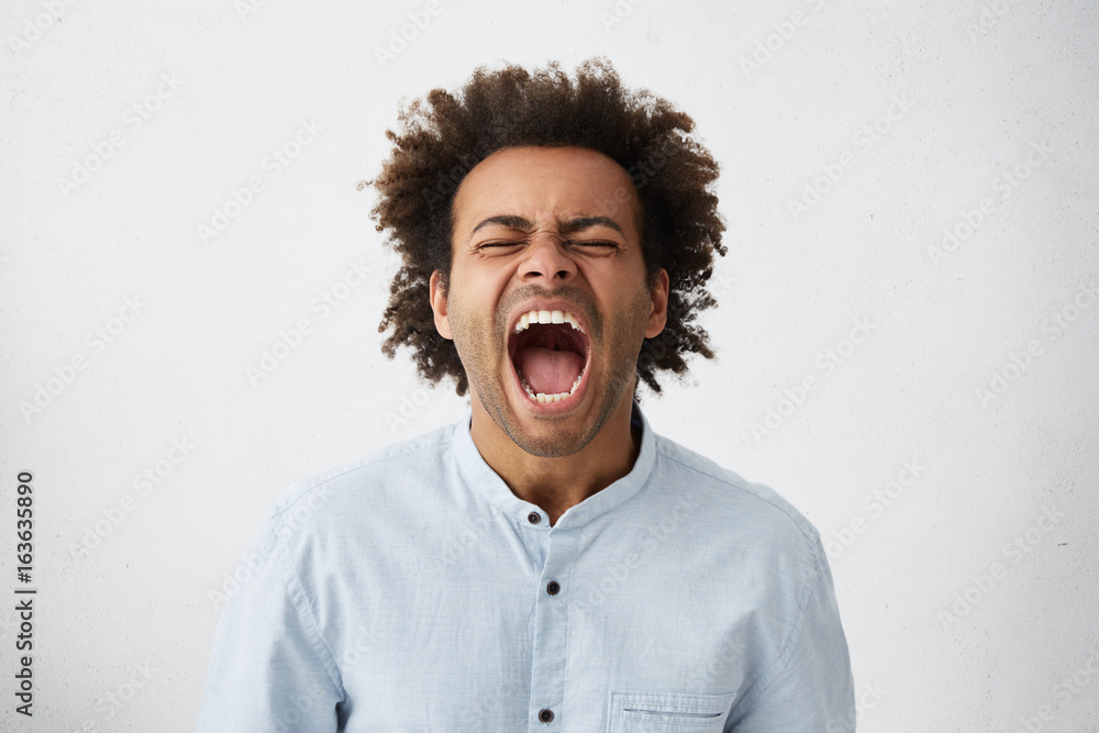 Portrait of dark-skinned African guy with curly hair screaming with wide opened mouth closing ...