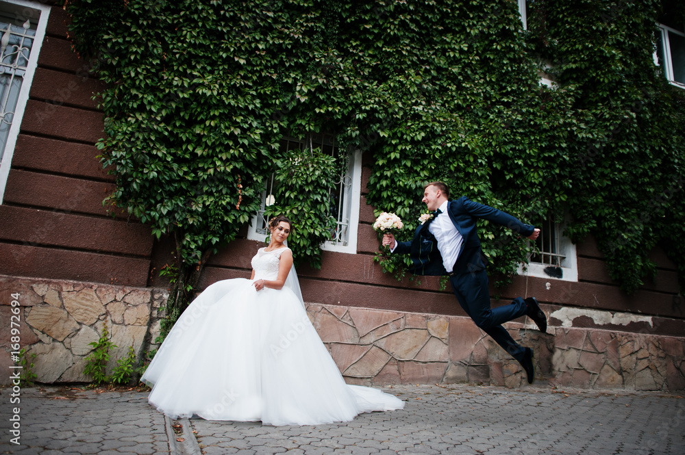 Elegant bride posing against the ivy wall while handsome groom jumping with a bouquet next to ...