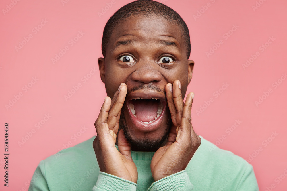 Stunned overjoyed African American male student screams with excitement, keeps hands near mouth ...