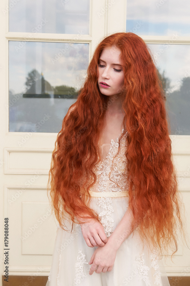 Beautiful red-haired girl with long curly hair in the bride, in a long ...