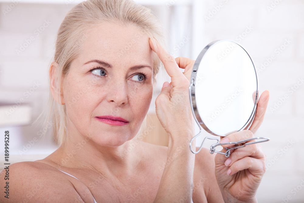 Middle aged woman looking at wrinkles in mirror. Plastic surgery and collagen injections. Makeup ...