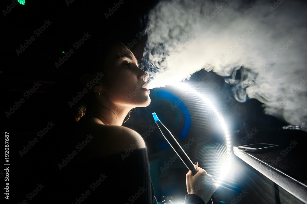 Young, beautiful girl smokes a hookah in the night club. Backlight LED ...