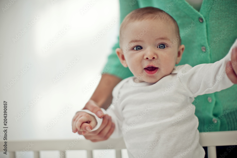 Cute smiling baby boy is supported by his mother's hands as she helps ...