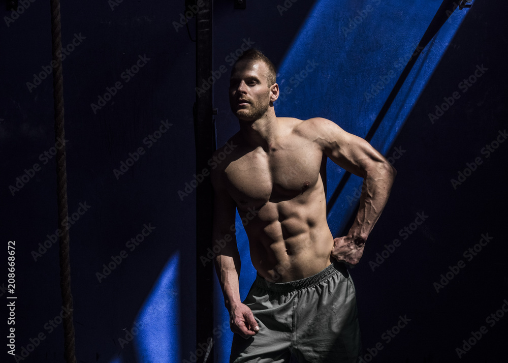 Attractive shirtless blond male bodybuilder in shorts indoors in dark ...