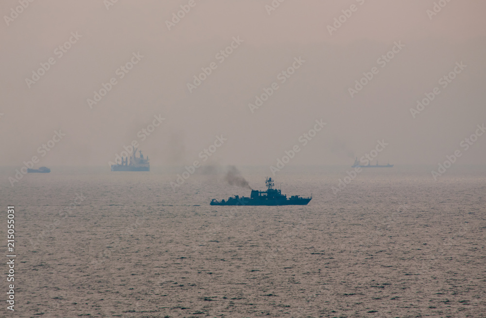 Military navy ship sailing among cargo ships on the horizon. Dark smoke ...