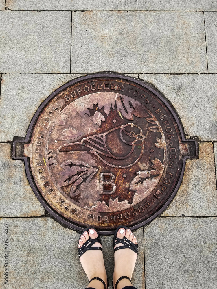 Girl's feet stand on sewer manhole on Vorobyovy Gory. Bird on hatch is ...