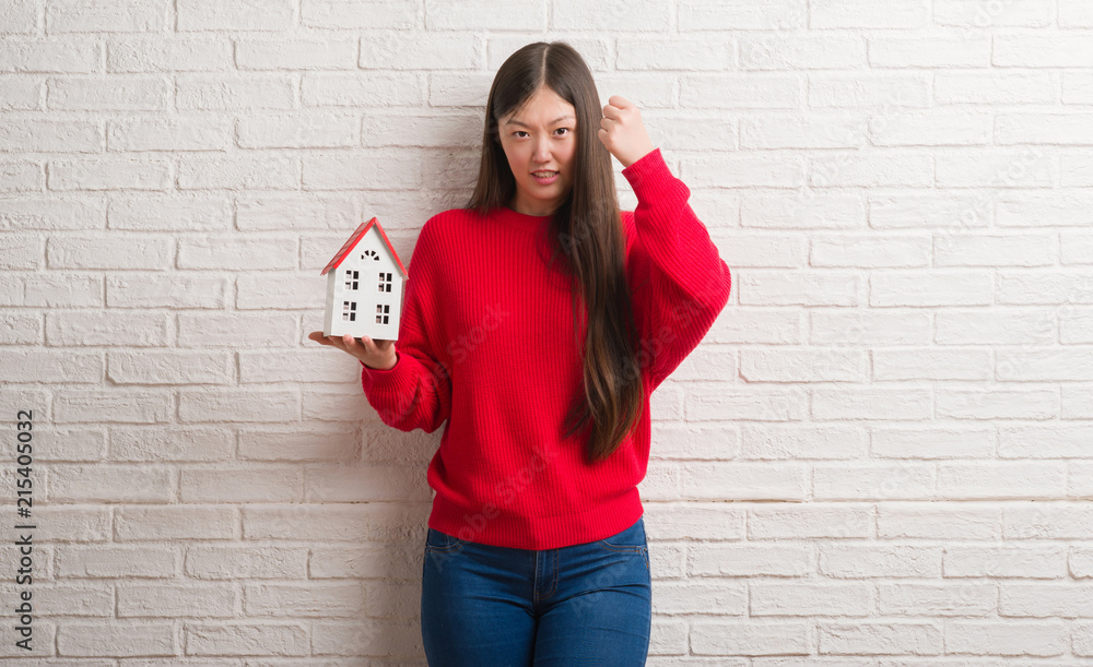 Young Chinese real state agent woman over brick wall holding house ...