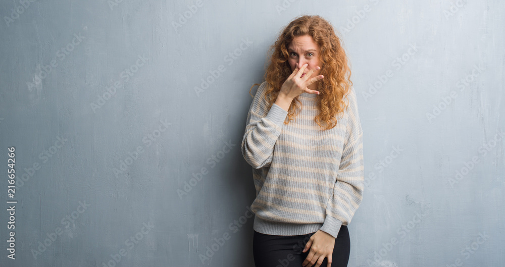 Young redhead woman over grey grunge wall smelling something stinky and ...