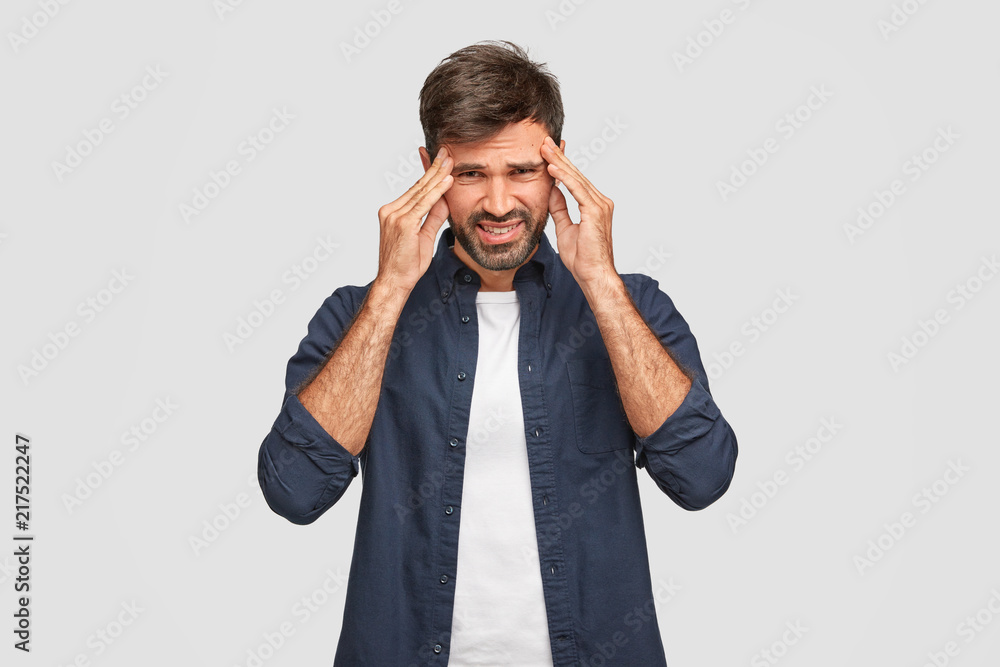 Handsome unshaven male keeps hands on temples, frowns face in ...