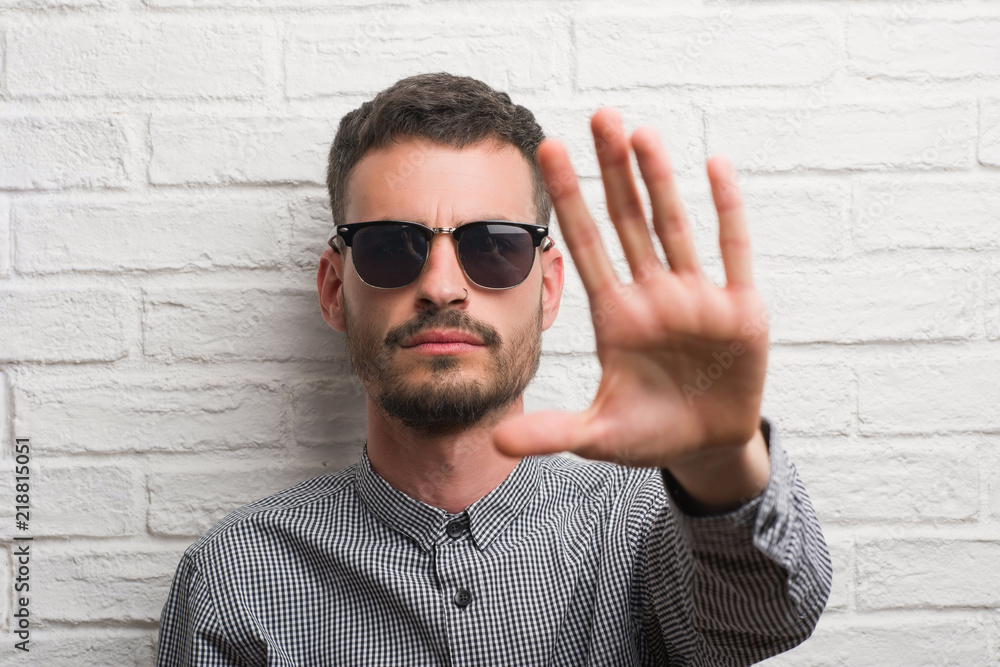 Young adult man wearing sunglasses standing over white brick wall with ...
