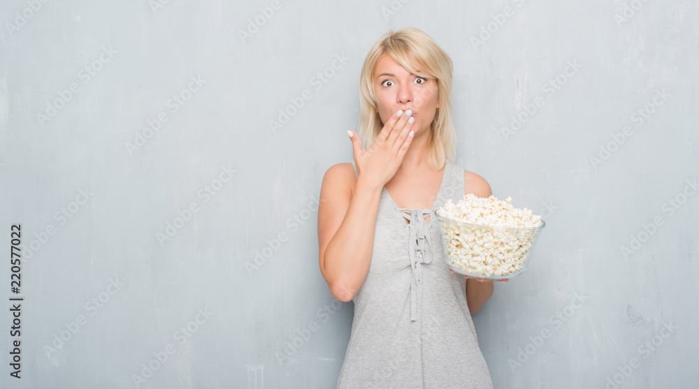 Adult caucasian woman over grunge grey wall eating pop corn cover mouth ...