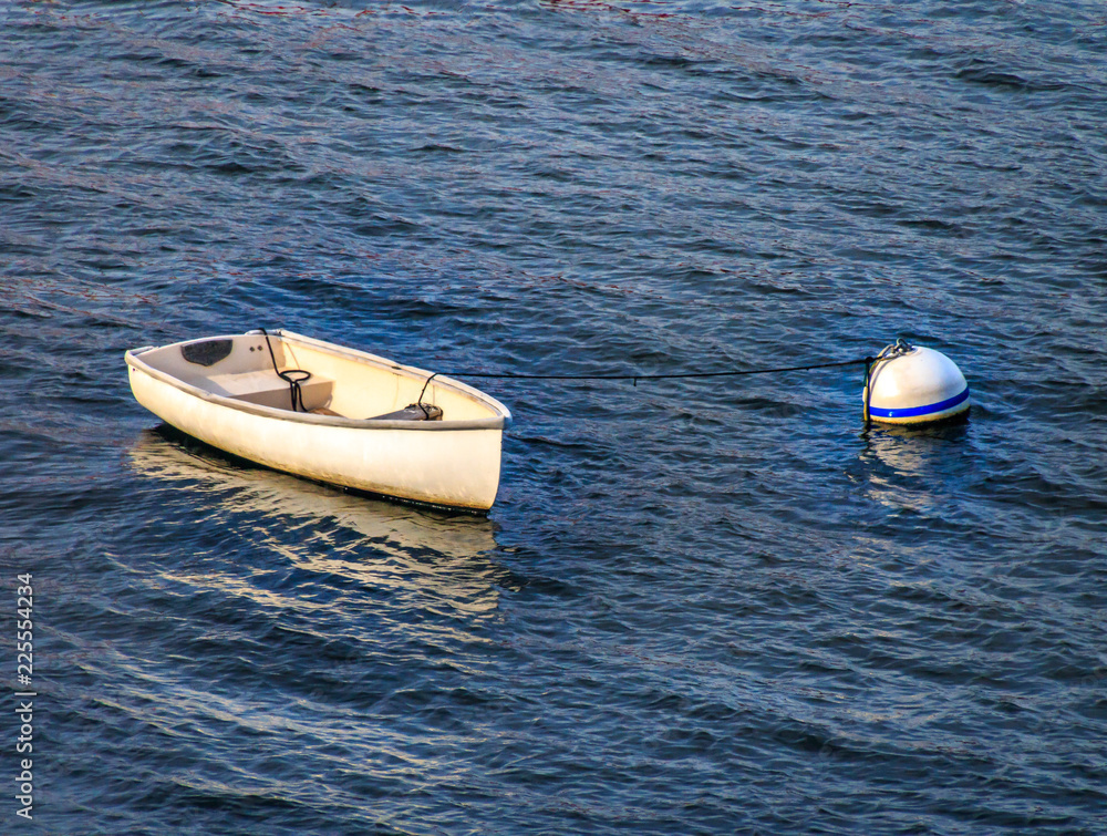 A small white rowboat skiff sits in the water attached by rope to a ...
