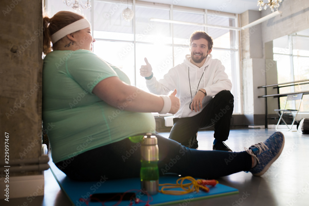 Full length portrait of obese young woman sitting on mat and talking to ...