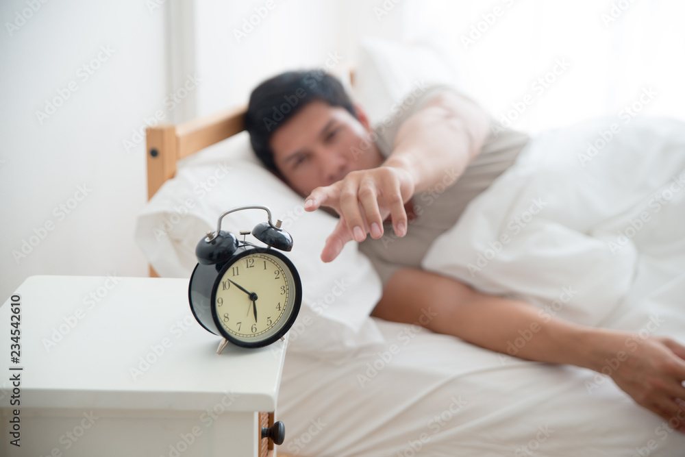Asian handsome man awakened by alarm clock in the bed at morning time ...