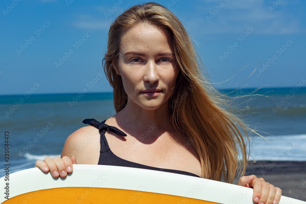 Surfer girl in bikini with surfboard posing on black sand beach. Face ...
