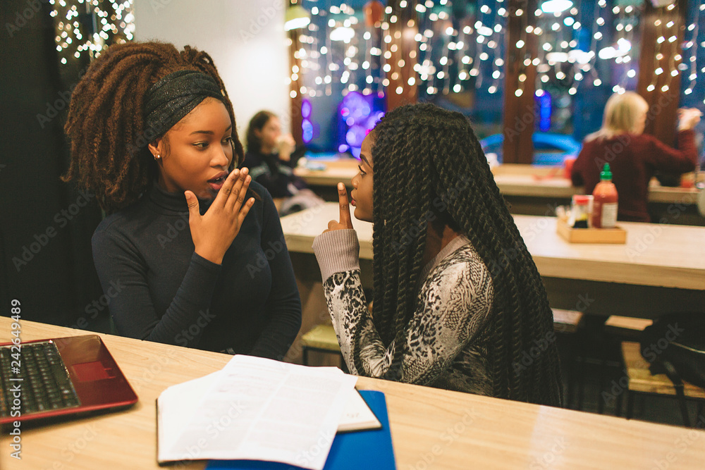 Two young african women gossip at table. On of them looks wondered ...