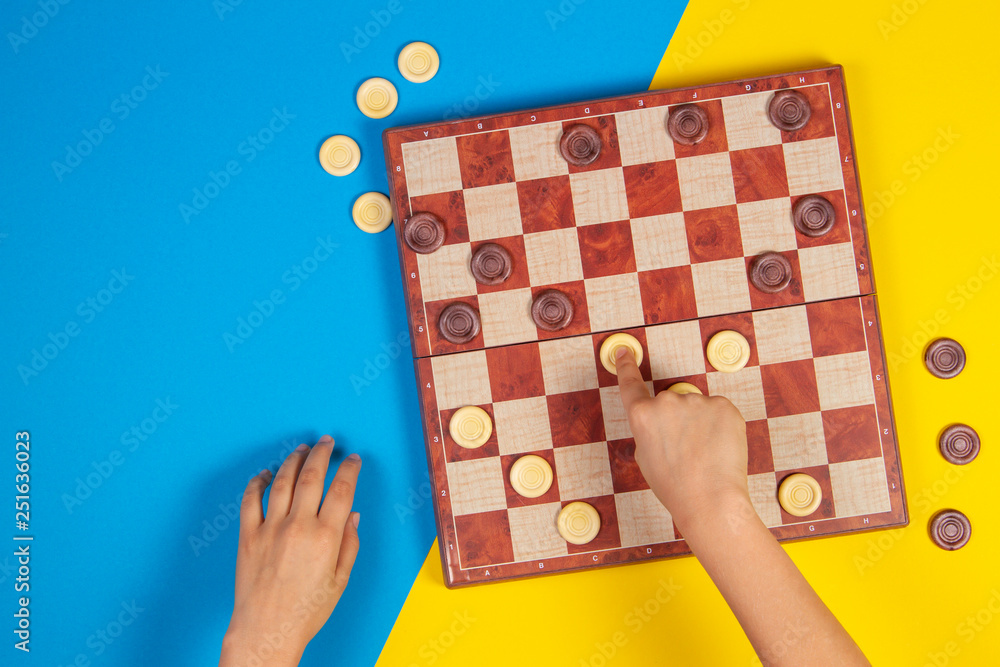 Child hands playing checkers on checker board game over yellow and blue ...