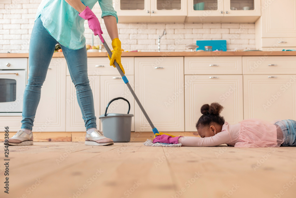 Funny curly girl lying on the floor while mother mopping the floor Stock Photo | Adobe Stock