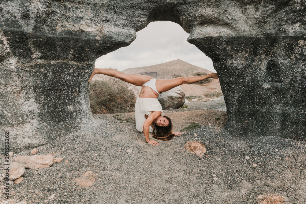 Pretty barefoot woman performing handstand with split in rough stone arch while exercising in ...