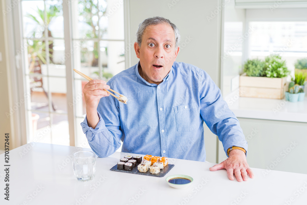 Handsome senior man eating take away sushi using chopsticks at home ...