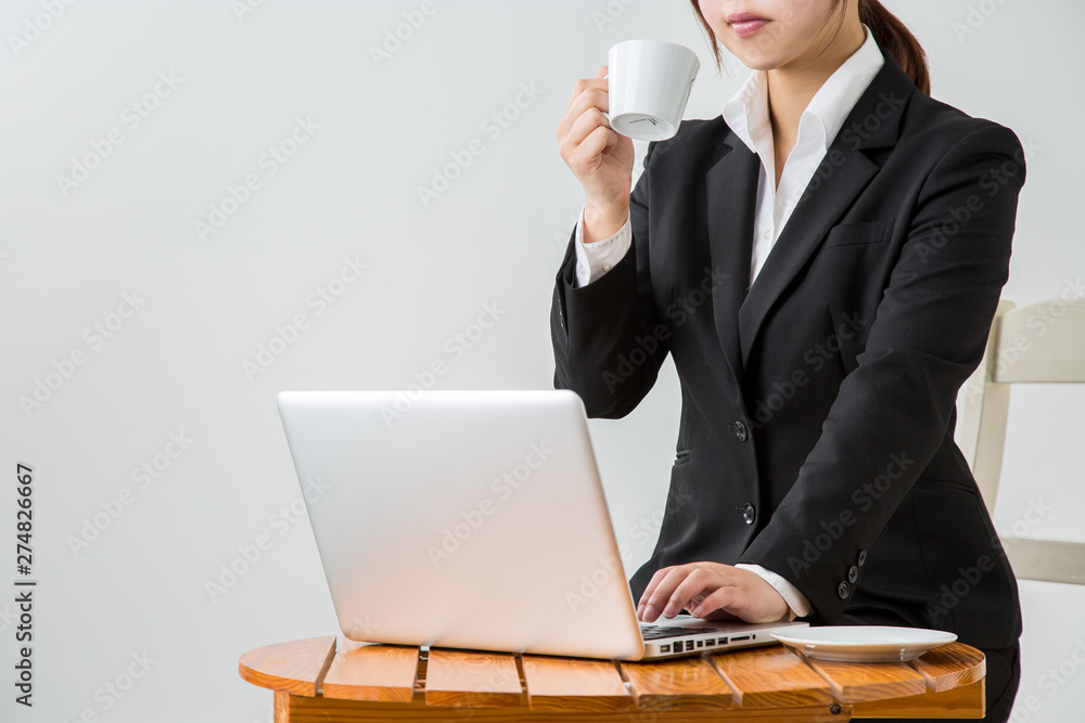 白背景でスーツをきた女性がノートパソコンで仕事しながらコーヒーを飲んでいる Stock Photo | Adobe Stock