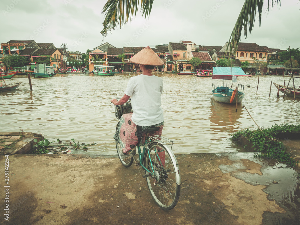 Woman with traditional vietnamese hat cycling on the river bank at Hoi An, famous travel ...