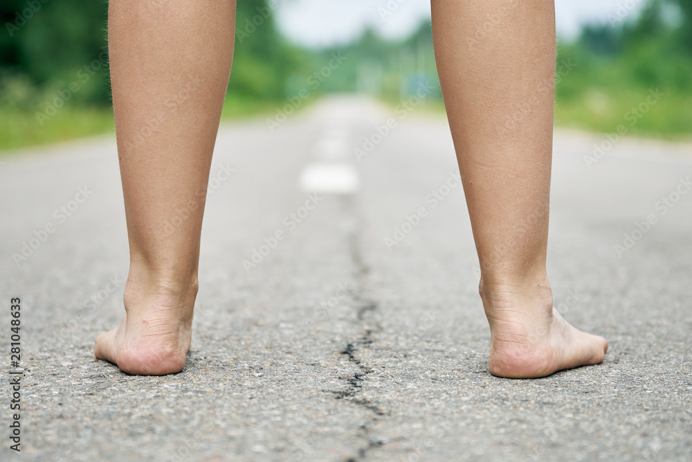Rear view of bare feet of a young girl standing on the asphalt road ...