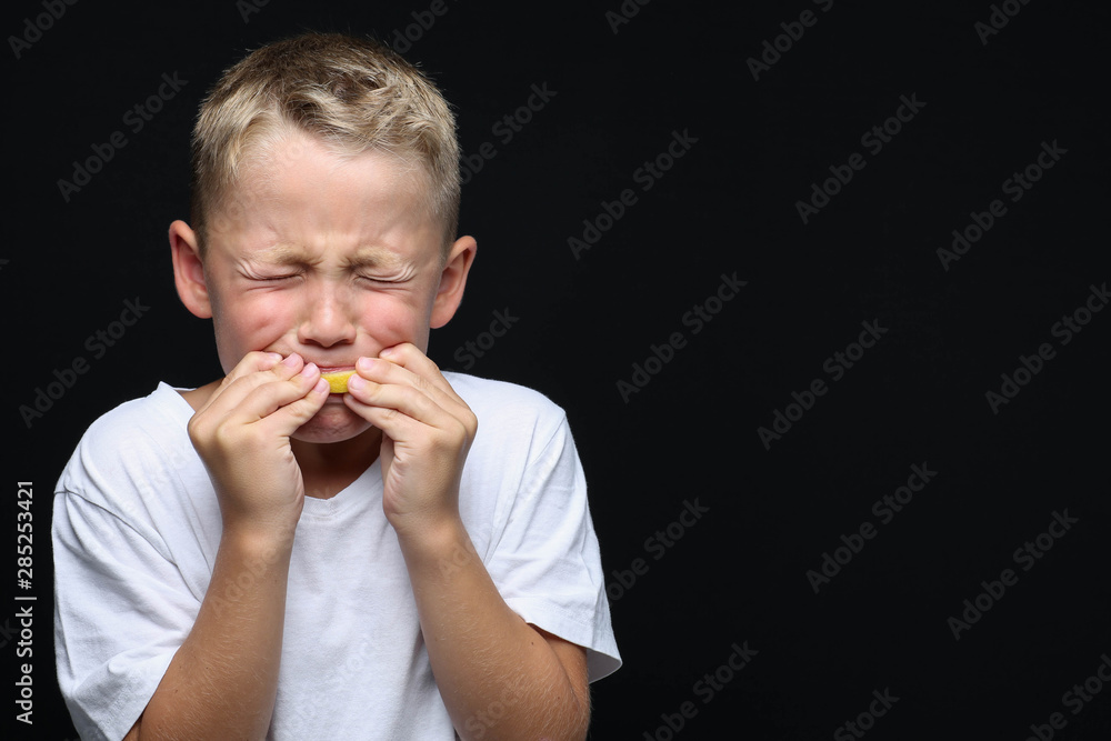 Little, blond boy is eating a piece of a lemon in front of black ...