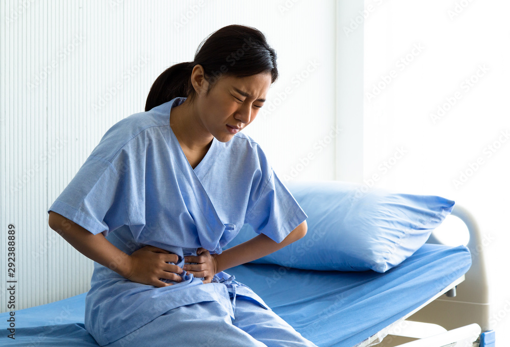 Young woman patient suffering from abdominal pain while sitting on bed ...