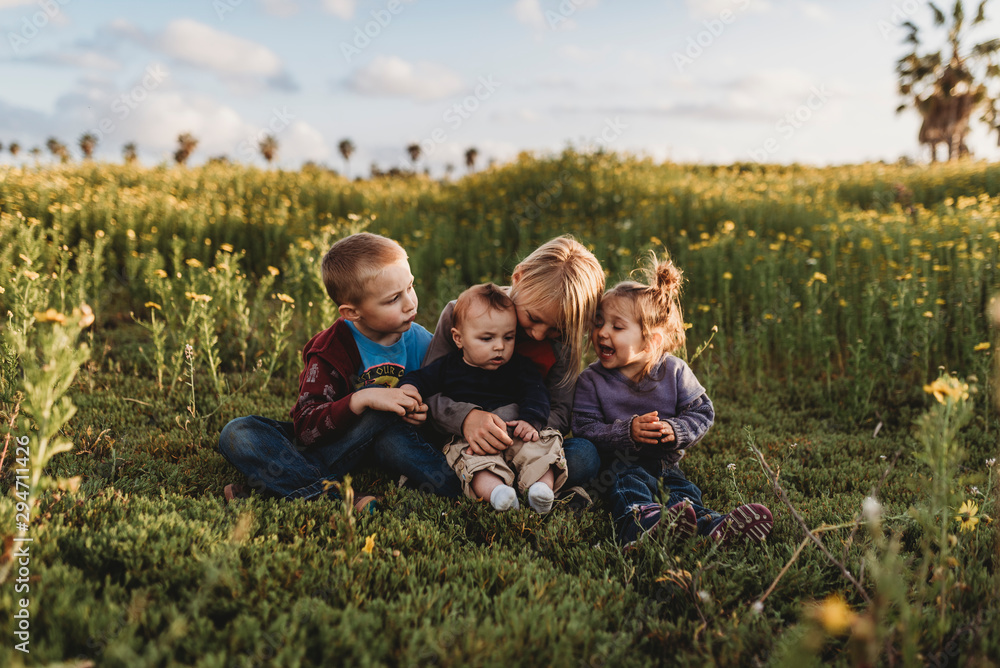 Four siblings smiling at each other in field of flowers with blue sky ...