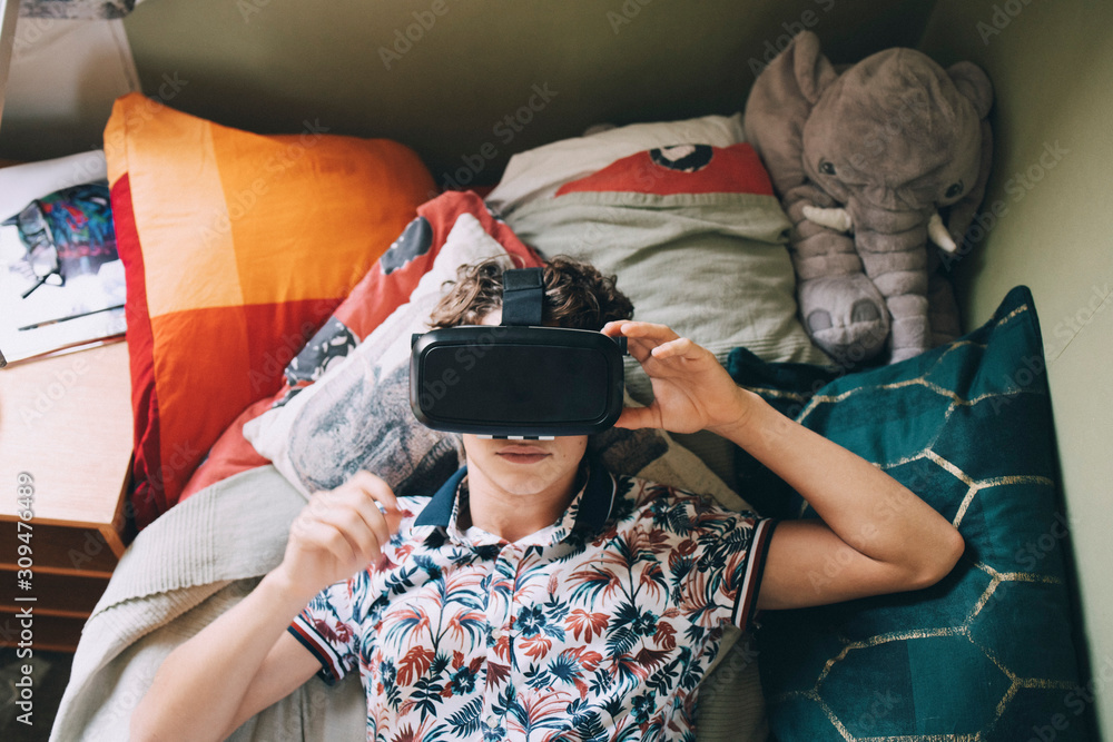 Teenage boy wearing virtual reality headset while lying on bed at home ...