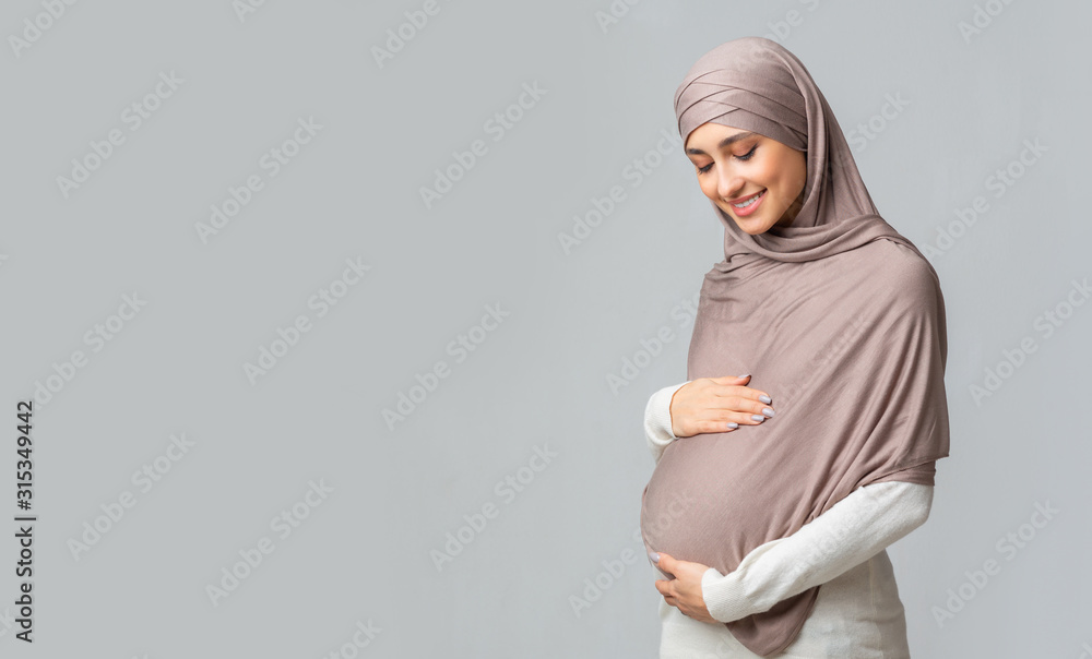 Pregnant muslim woman embracing her belly, posing over grey background ...