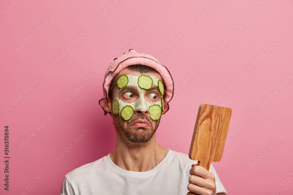 Impressed young man stares at wooden mirror, applies clay mask and ...