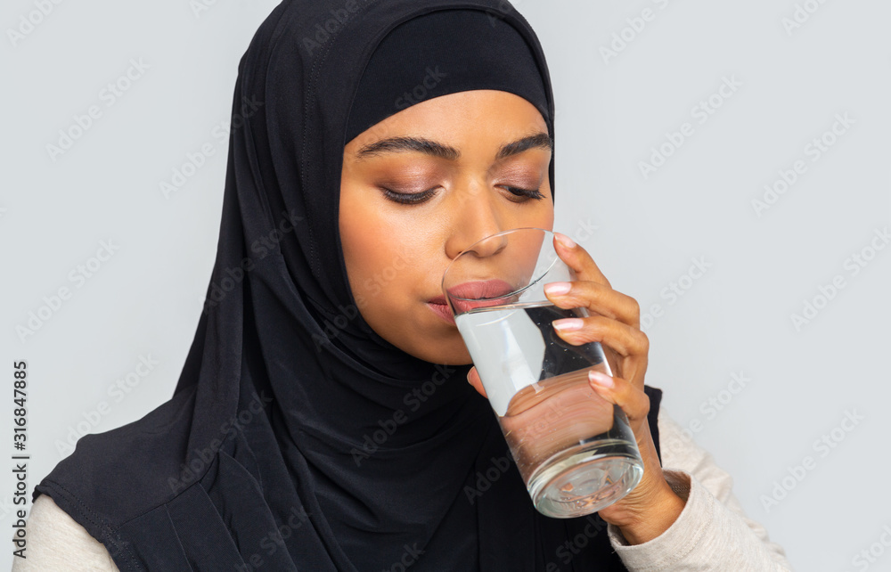 Thirsty black muslim woman in hijab drinking mineral water from glass ...