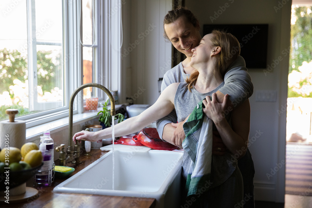 straight caucasian millennials couple in the kitchen kissing and washing dishes in the morning ...