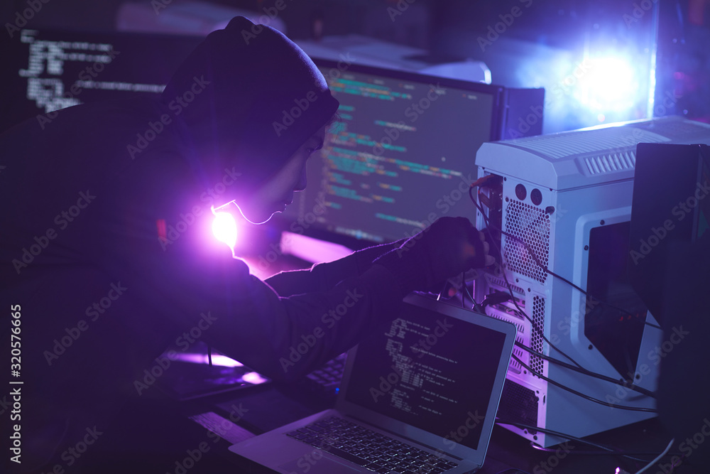 Side view at unrecognizable cyber security hacker wearing hood while working on computer system ...