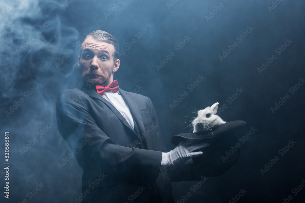 emotional shocked magician holding white rabbit in hat, dark room with ...