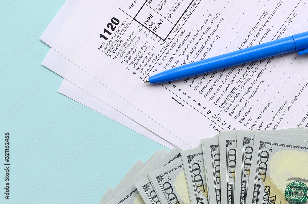 1120 tax form lies near hundred dollar bills and blue pen on a light ...