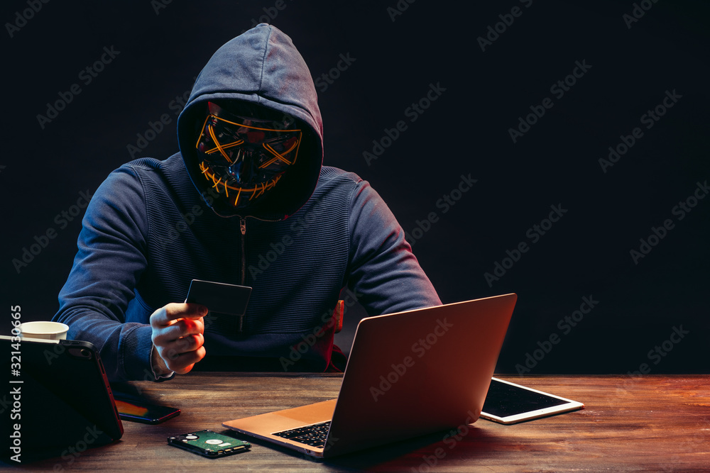 aggressive anonymous hacker man in mask sit with laptop isolated in dark space, man uses ...