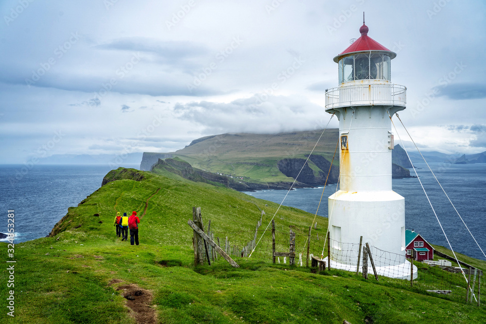 The lighthouse on Mykines island in Faroe Islands. Three hikers hiking ...