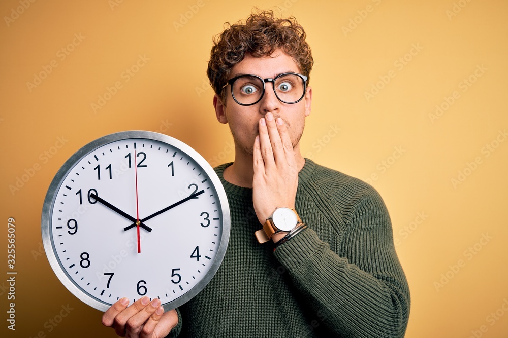 Young blond man with curly hair wearing glasses holding big clock over ...