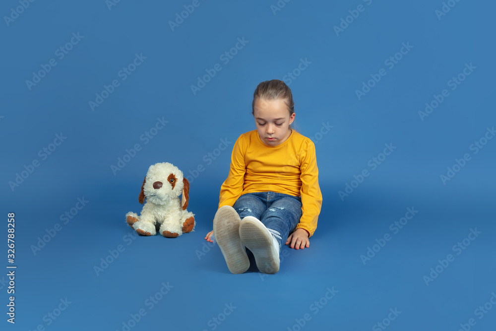 Portrait of sad little girl sitting isolated on blue studio background ...