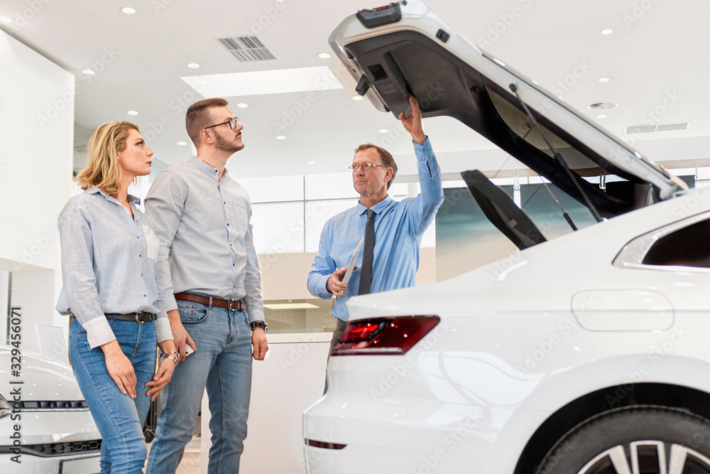 Sales manager helping young couple choose a new car by showing the size ...